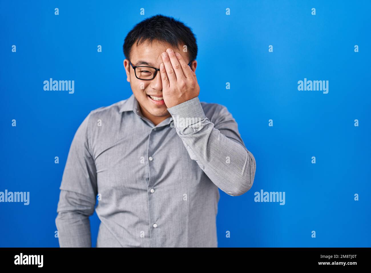 Young chinese man standing over blue background covering one eye with ...