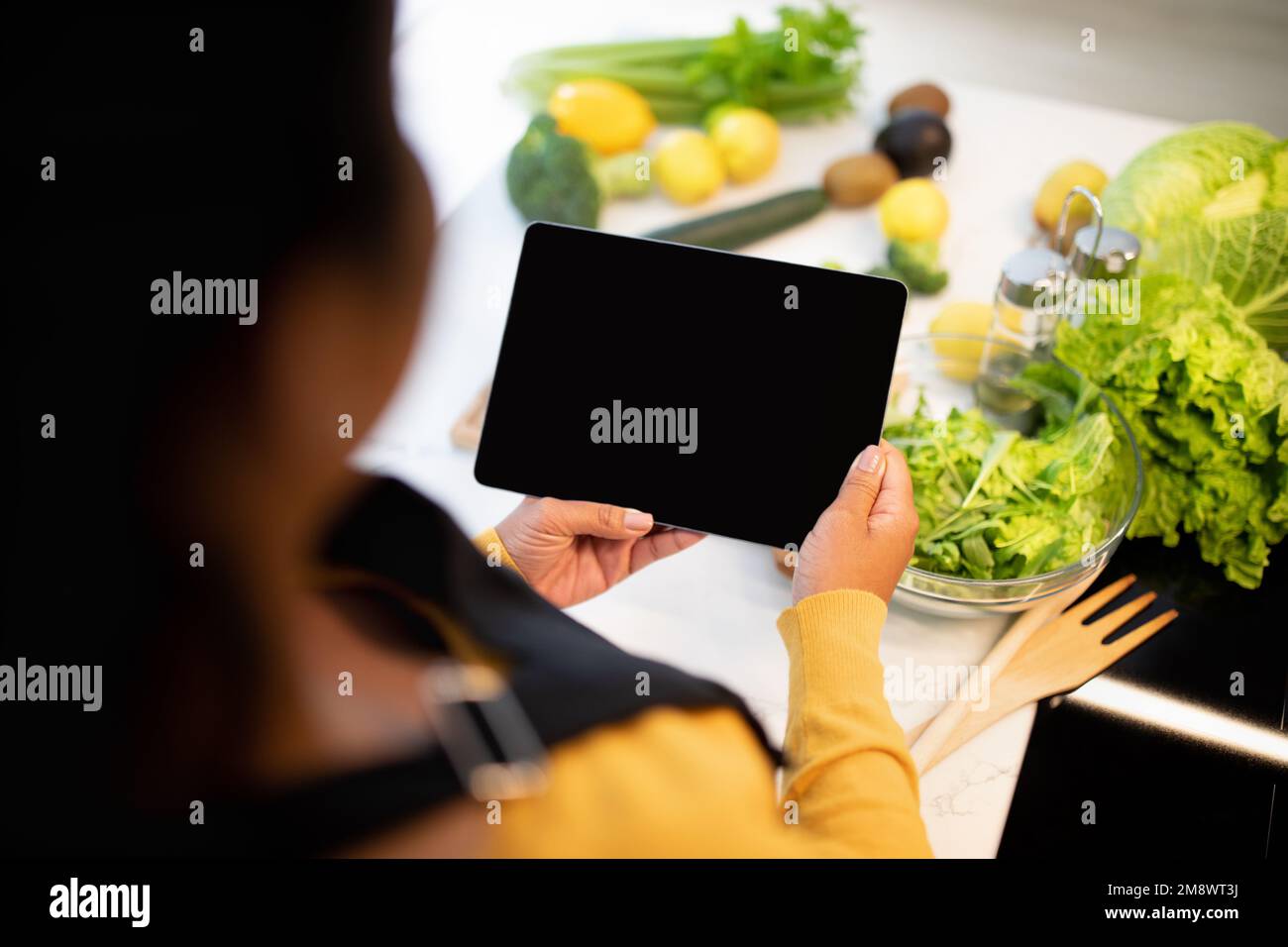 Young african american chef lady in apron cuts salad, use tablet with ...