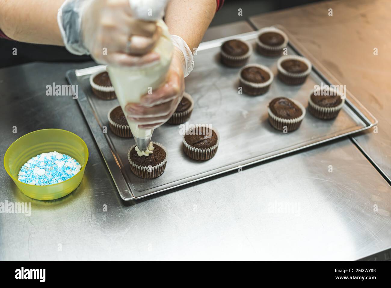 Unrecognisable baker piping white frosting on chocolate cupcakes on ...