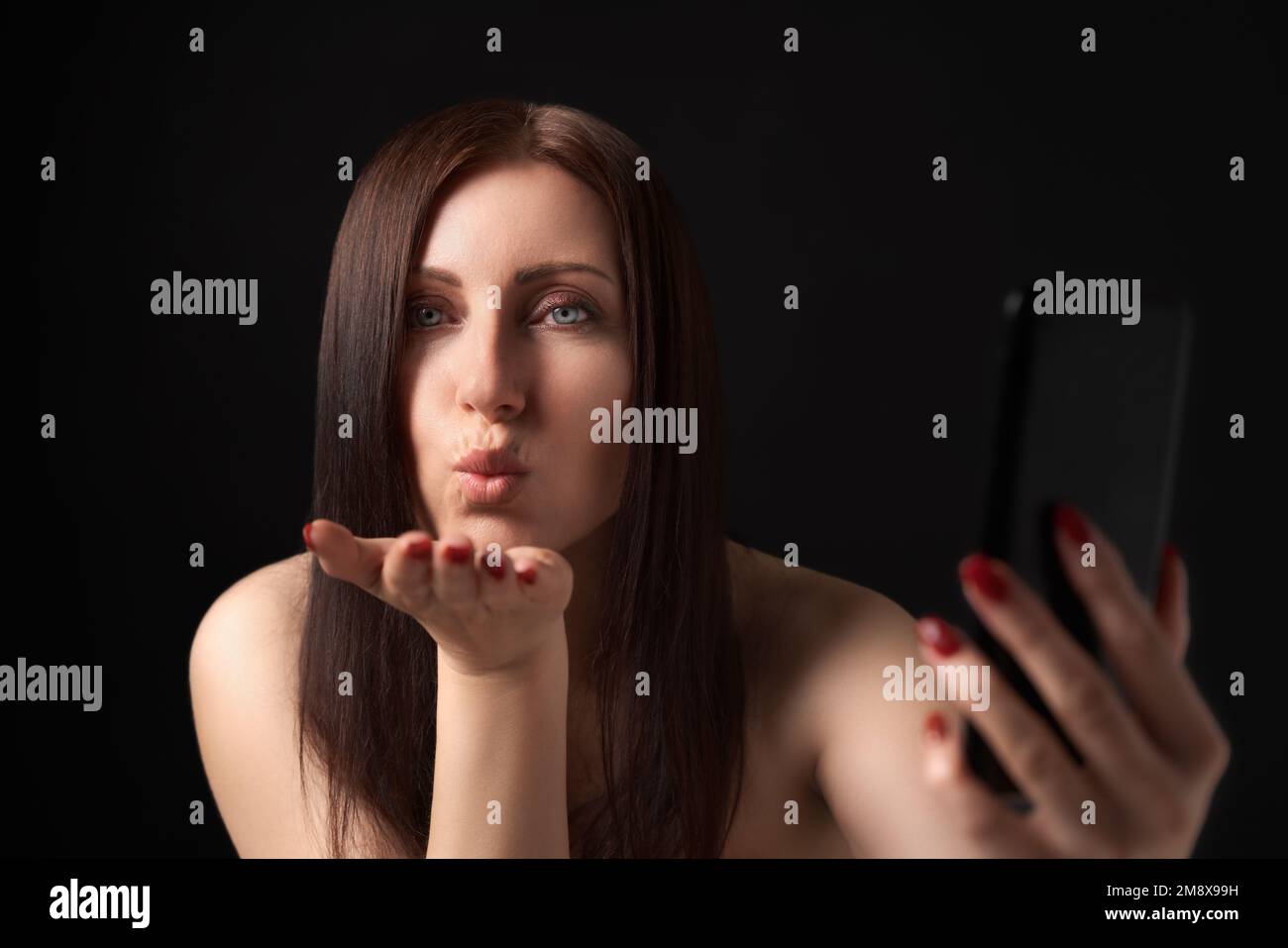 Woman blowing lips sending air kisses over palm and taking selfie to mobile phone camera. 40s ...