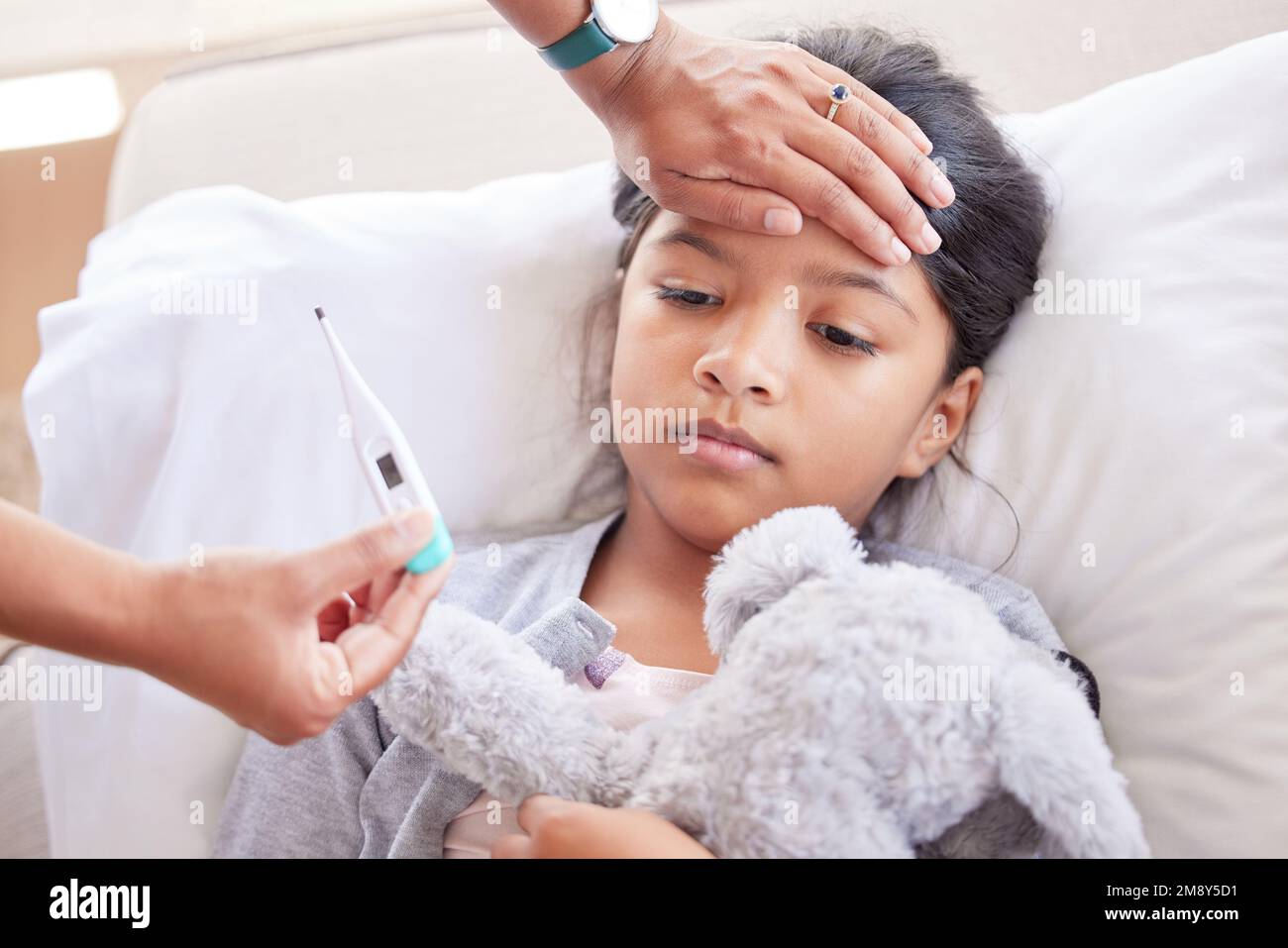 Little girl having her temperature taken with a thermometer by her mother at home. Small sick ...