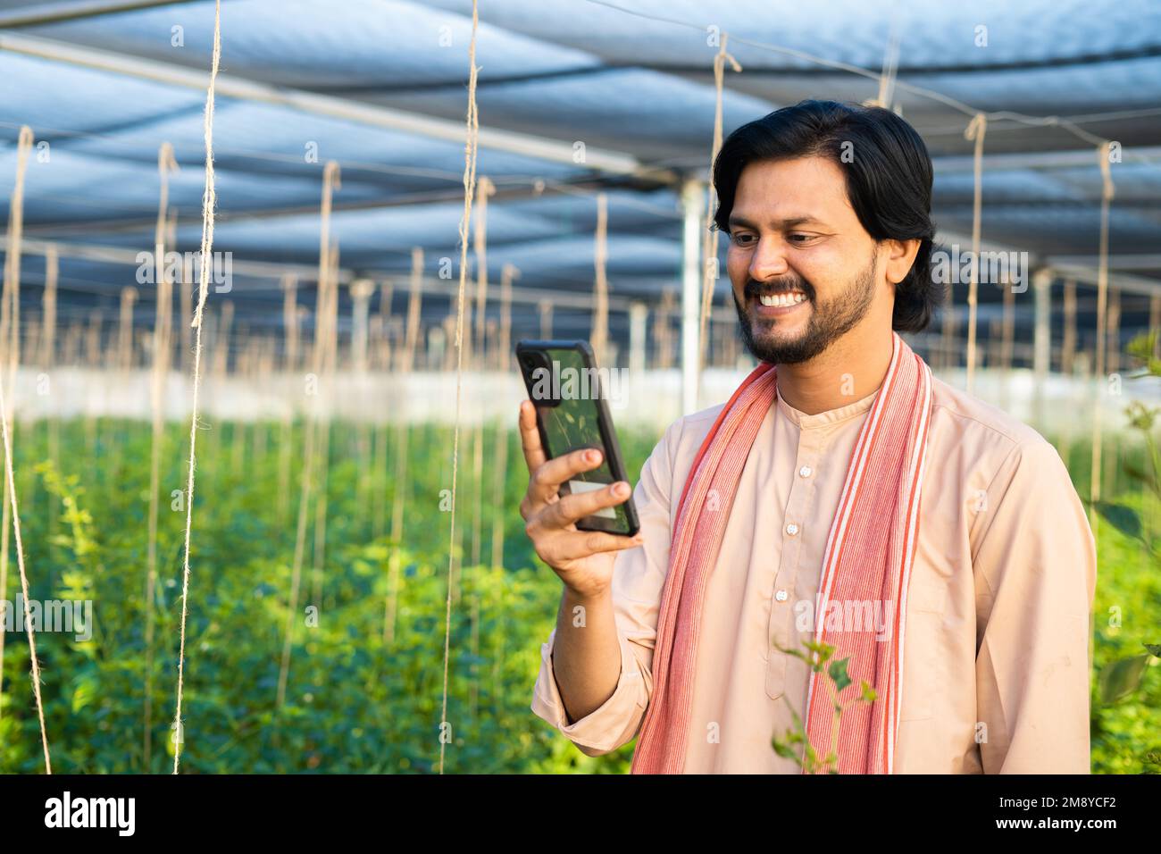 Happy smiling young farmer busy using mobile phone at greenhouse ...