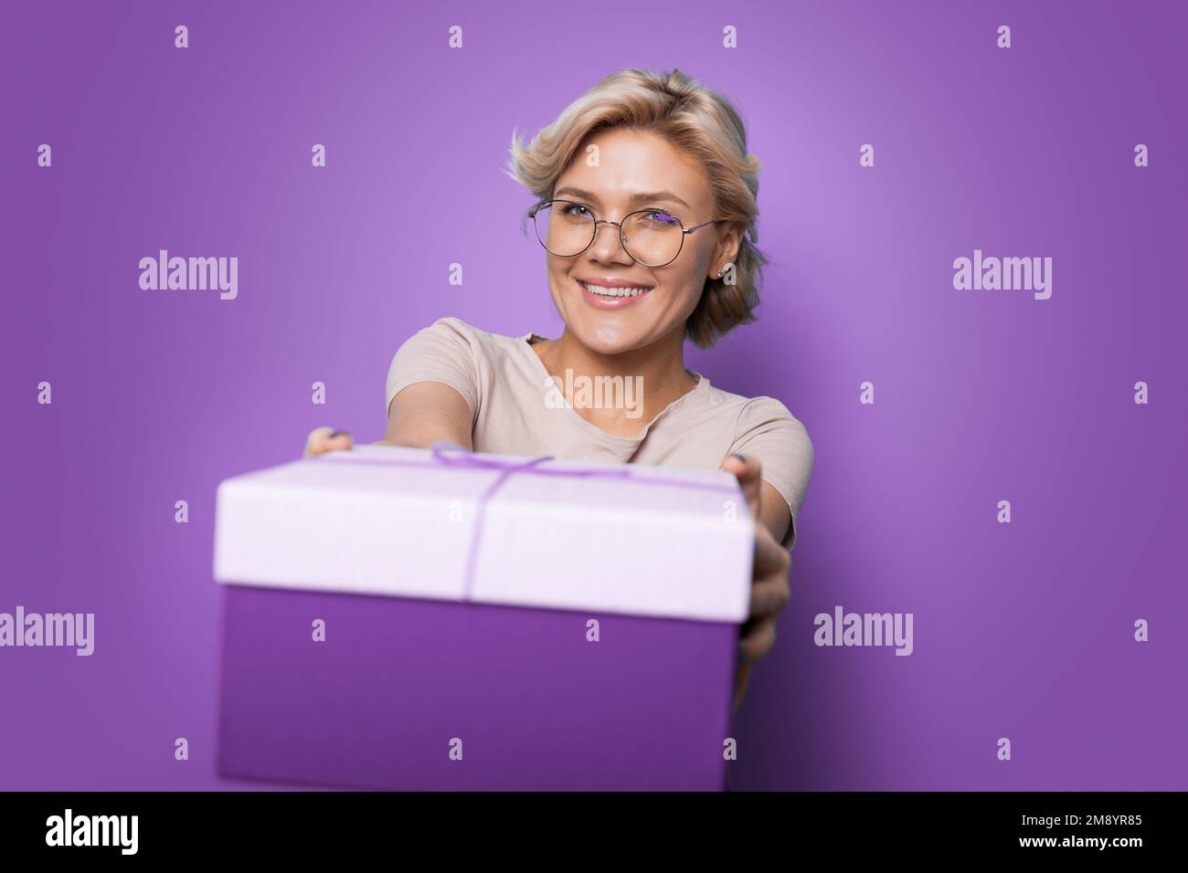 Beautiful young woman giving gift box at camera on purple studio ...