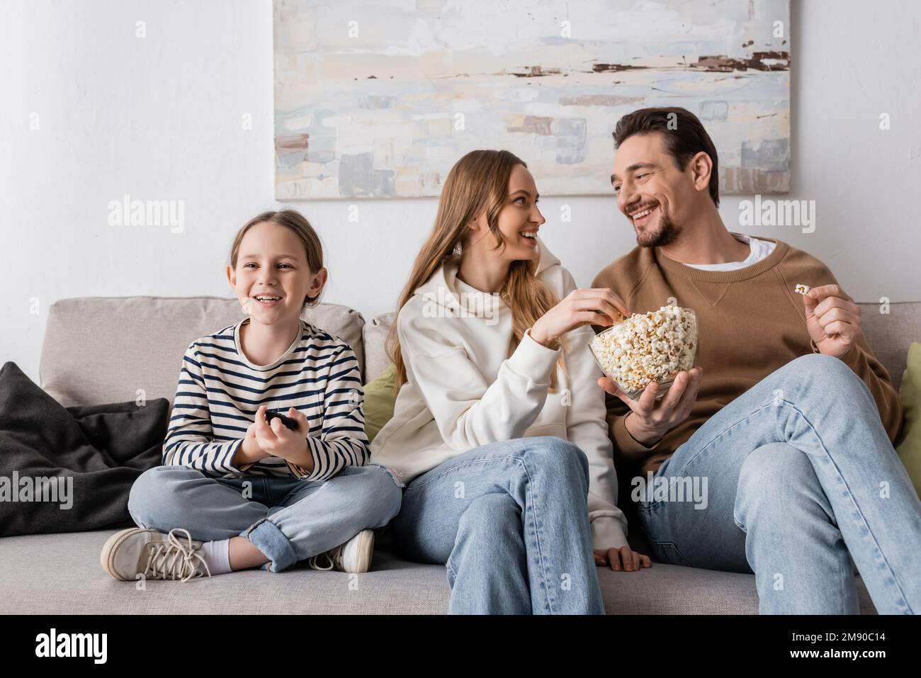 cheerful parents eating popcorn near happy daughter with remote ...