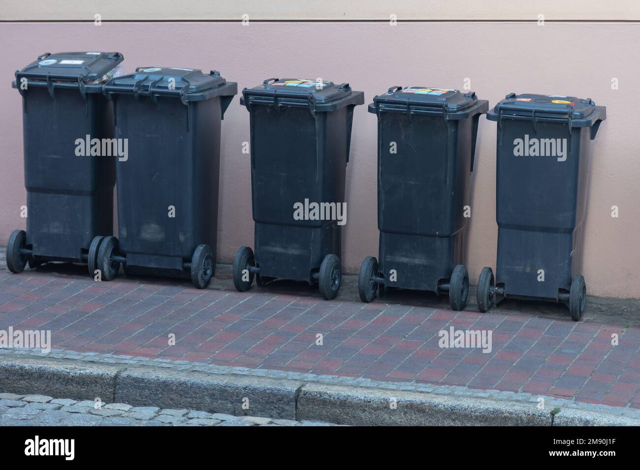 Black garbage cans standing in a row on a steeply sloping sidewalk ...
