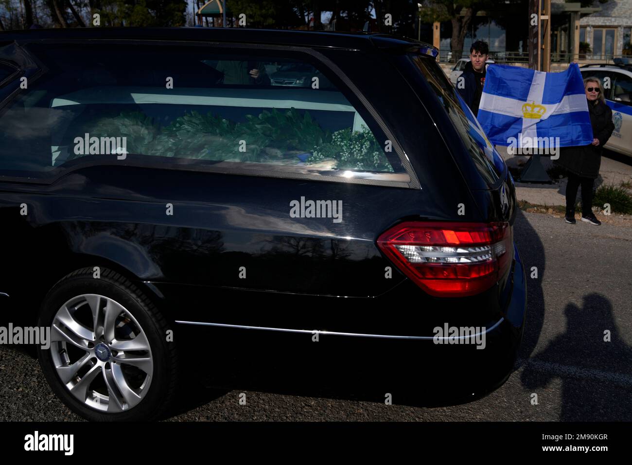 People hold a Greek flag with the crown as a hearse carrying the casket ...
