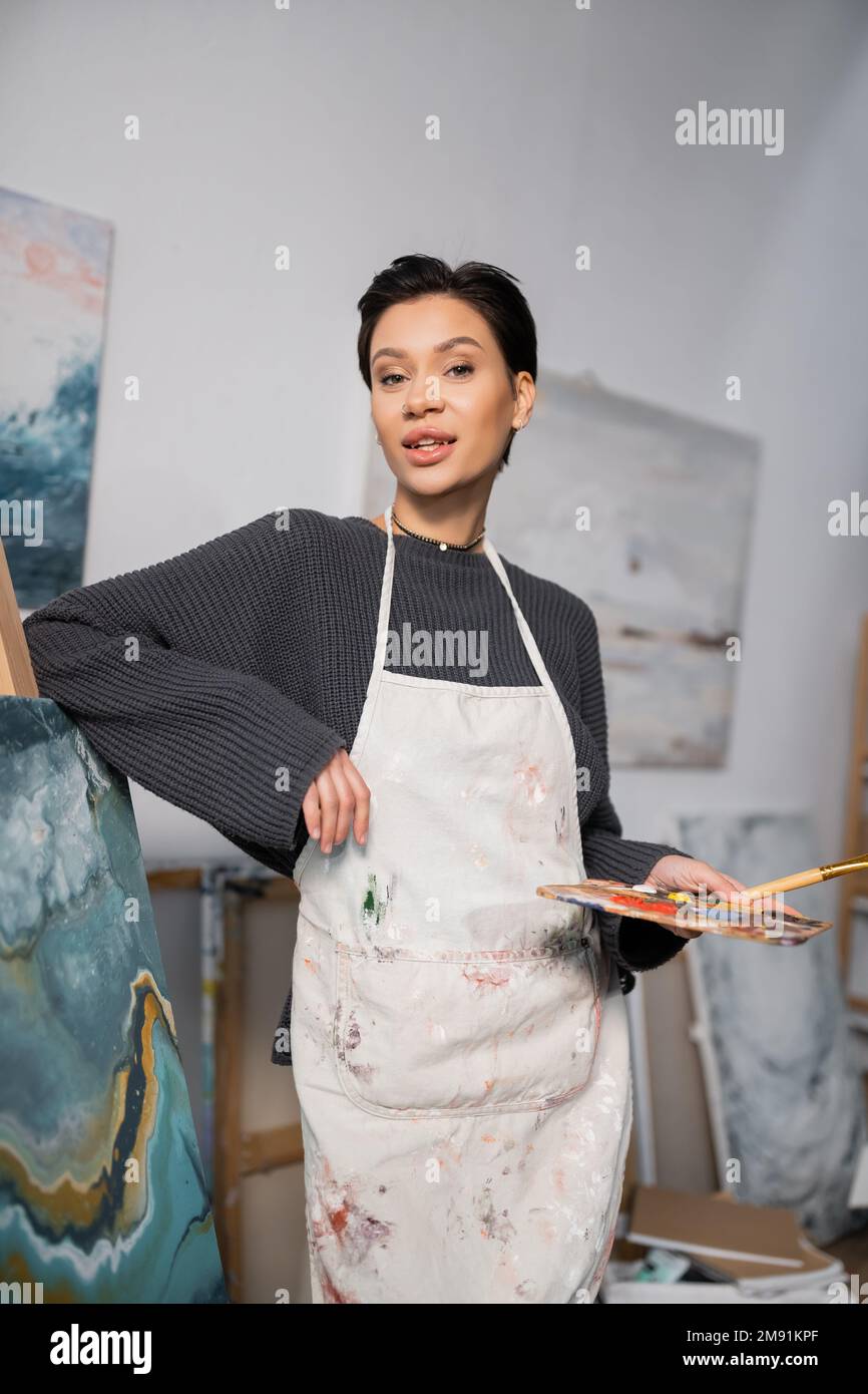 Brunette artist in dirty apron holding palette near drawing in workshop ...