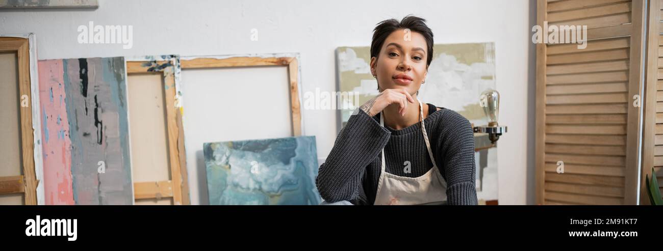 Brunette artist looking at camera near paintings in studio, banner ...