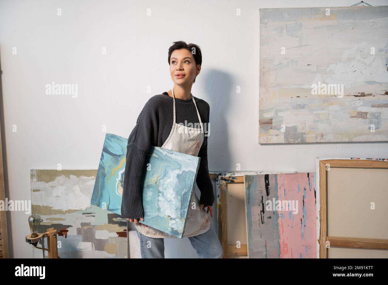 Dreamy artist in apron looking away near paintings in studio Stock ...