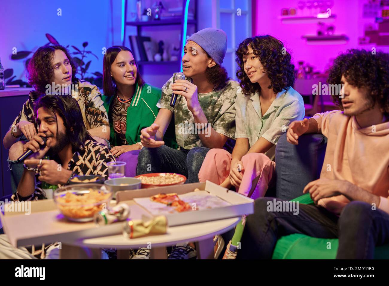 Young restful guys and girls sitting by served table in front of tv set ...