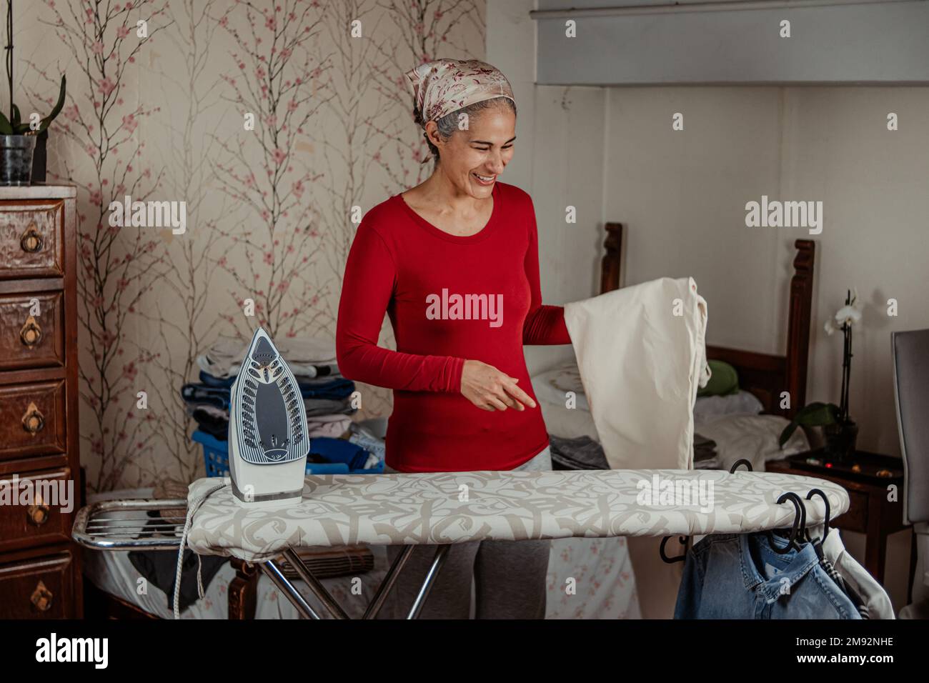 Cheerful mature female in domestic clothes smiling and ironing casual ...
