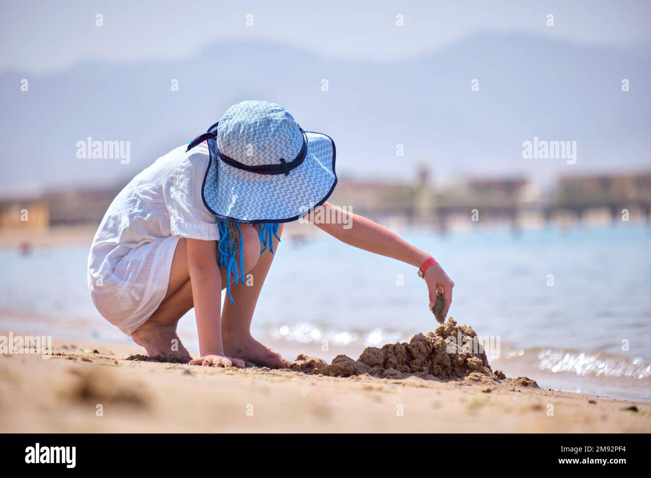 Pretty child girl in big hat and white dress playing with wet sand on ...