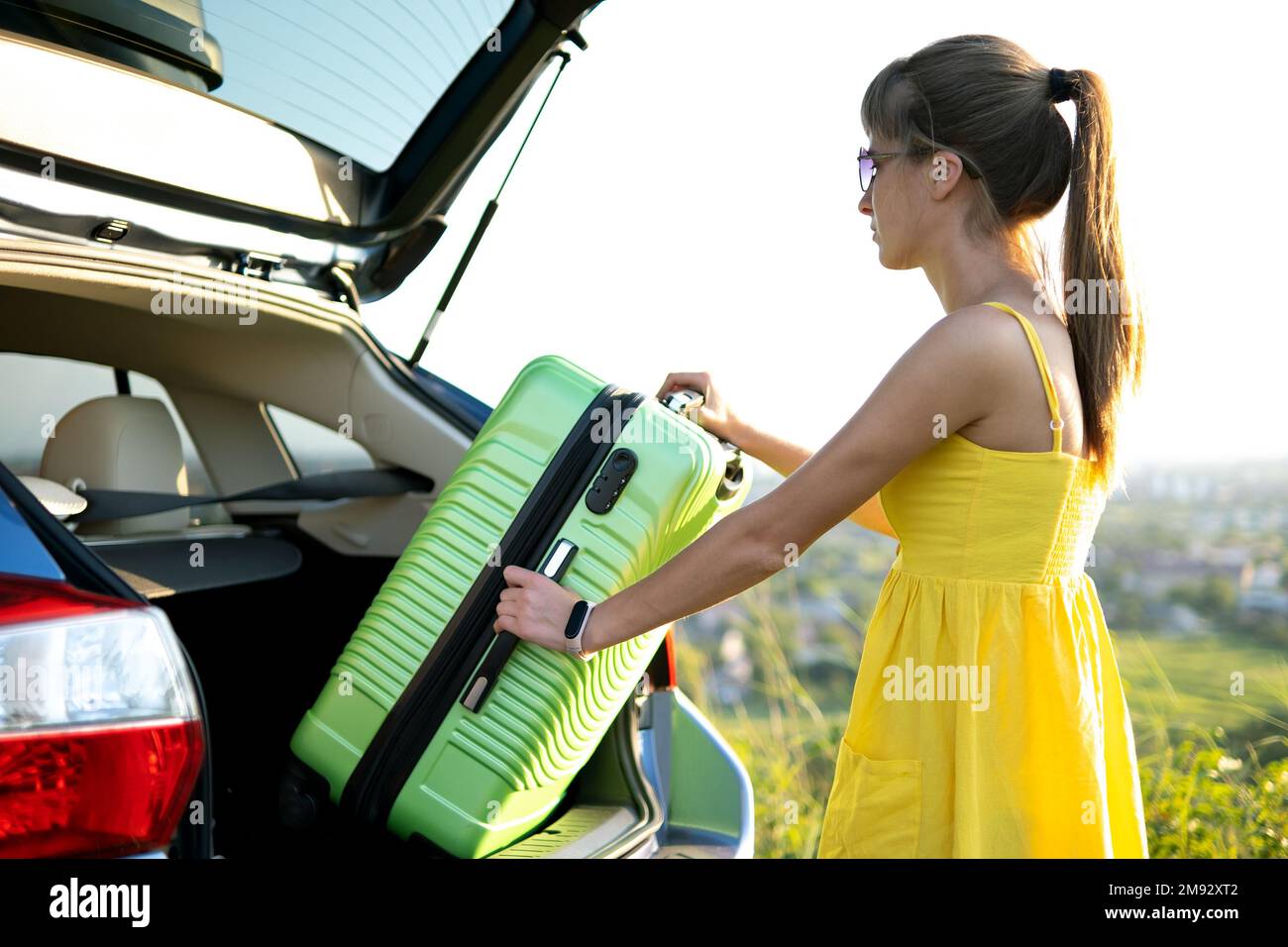 Young female driver putting green suitcase inside her car trunk. Travel ...