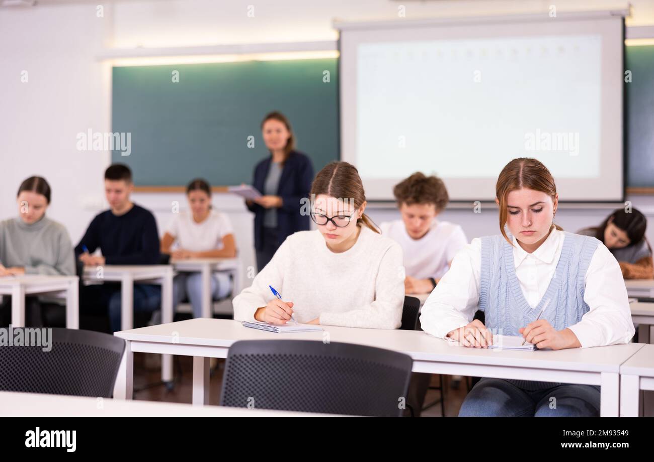 Group of school teenagers with pens and notebooks studying in classroom ...