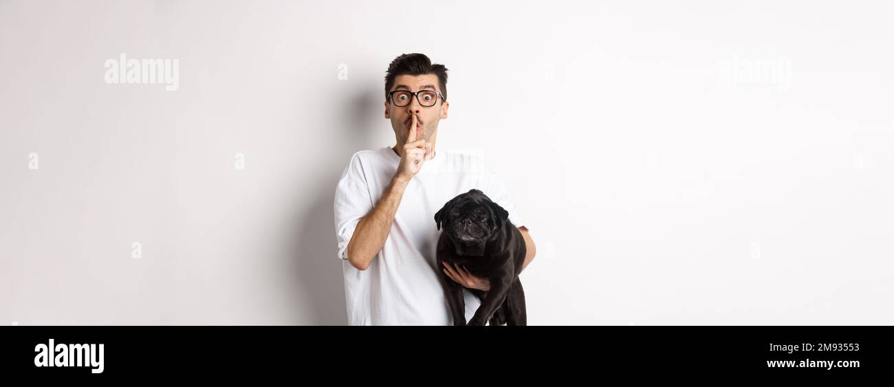 Image of funny young man holding cute black pug dog and shushing at ...