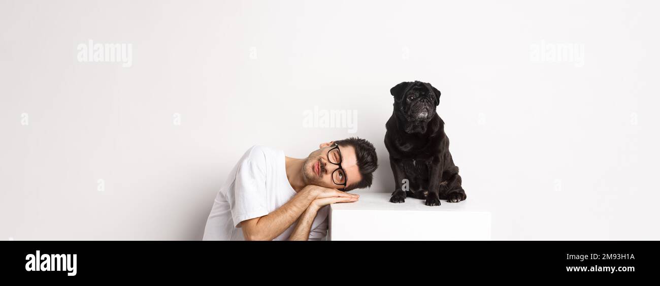 Handsome young man lay head near cute black pug, smiling and looking up ...