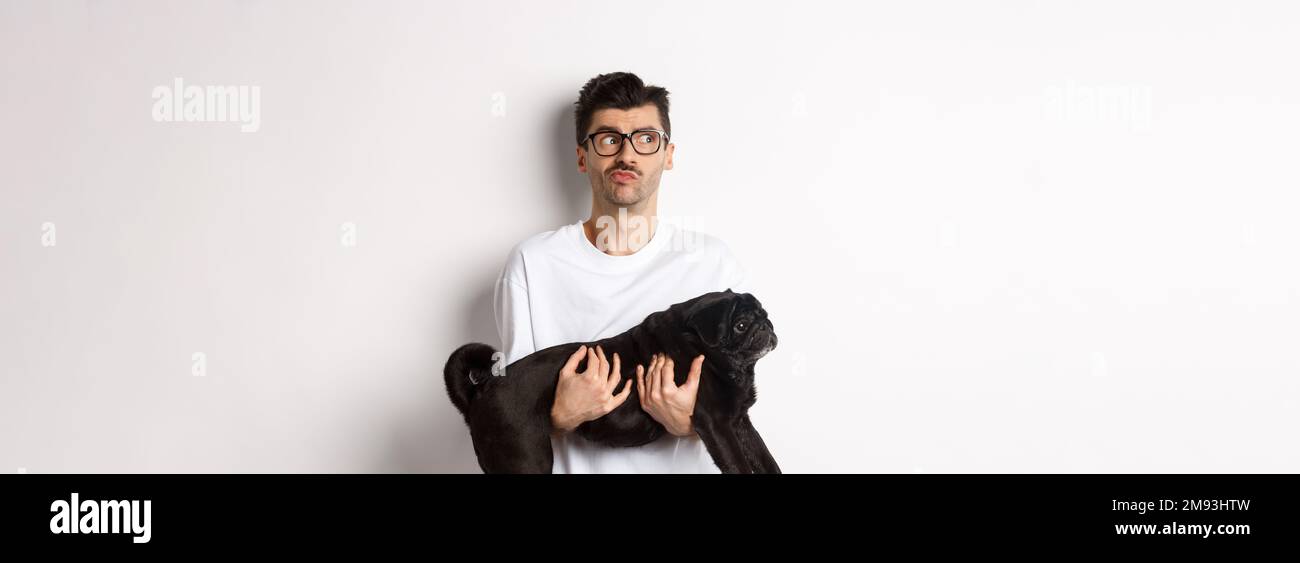 Confused hipster guy holding black cute pug and looking left, feeling ...