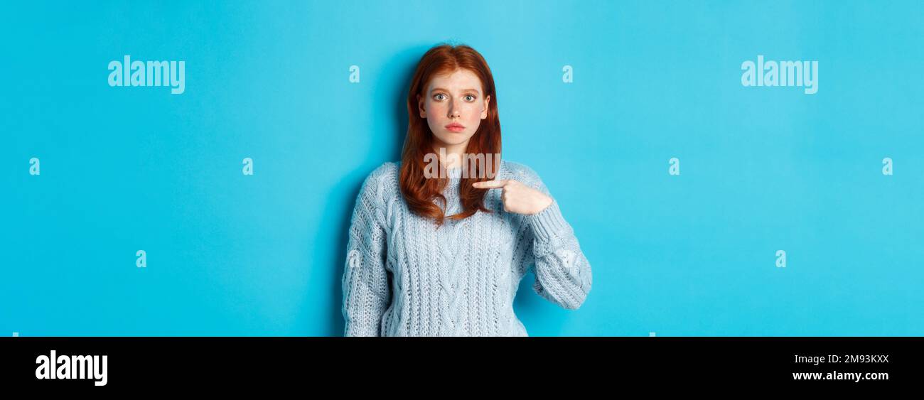 Nervous and confused redhead girl pointing at herself, standing in ...
