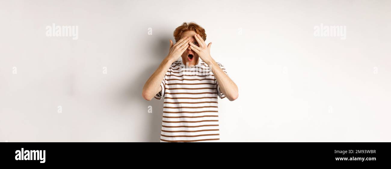 Shocked and embarrassed redhead guy covering eyes, peeking through ...