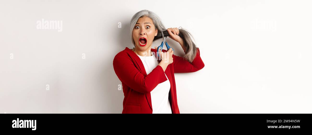 Scared asian senior woman cutting hair with scissors and screaming in ...
