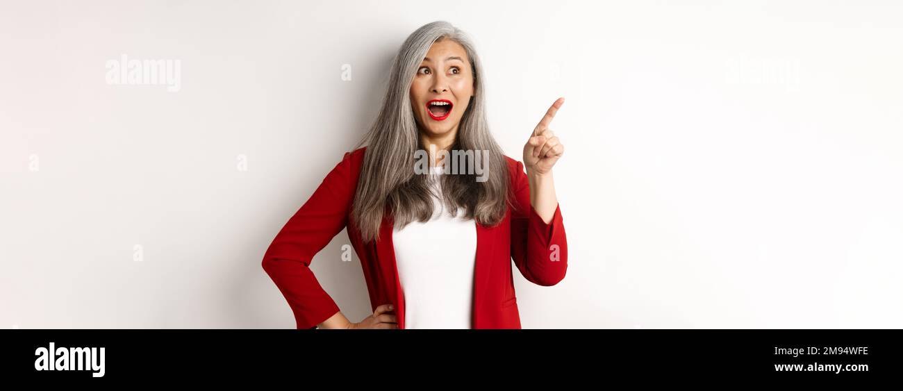 Surprised senior asian lady pointing upper left corner, checking out ...