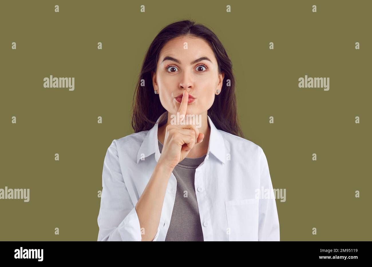 Portrait of young woman on khaki studio background holds finger at lips make shush silence sign ...