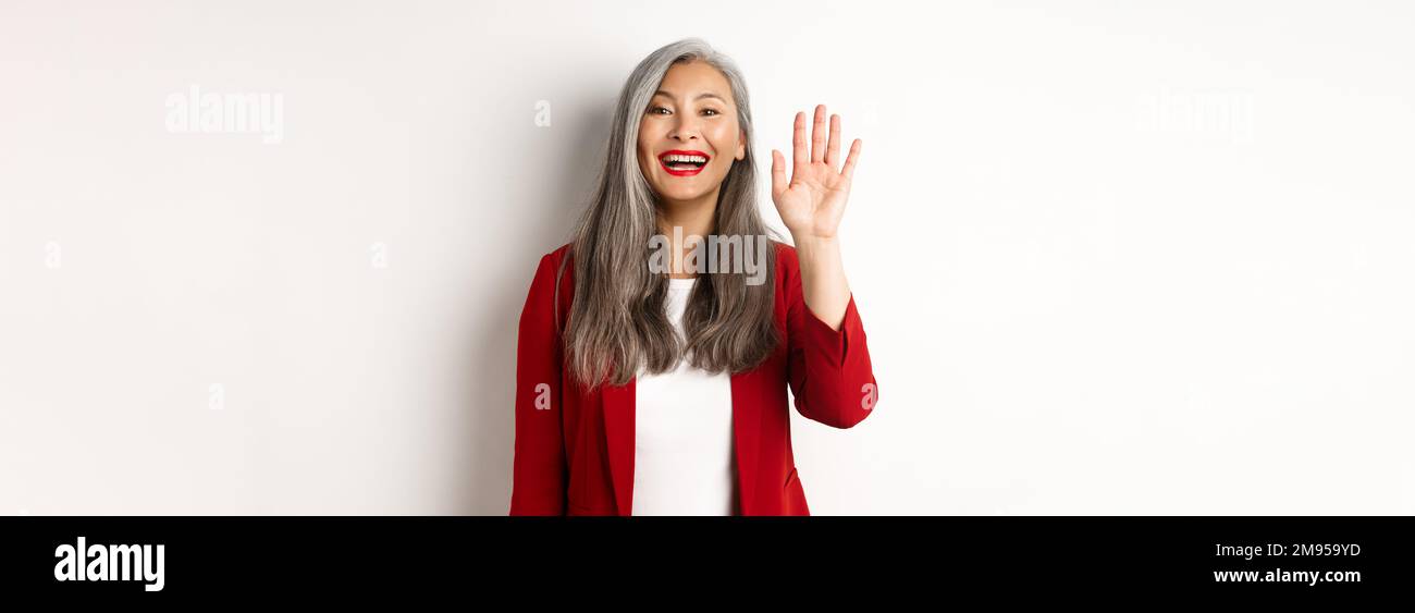 Friendly asian senior lady in elegant blazer saying hi, waving hand and ...