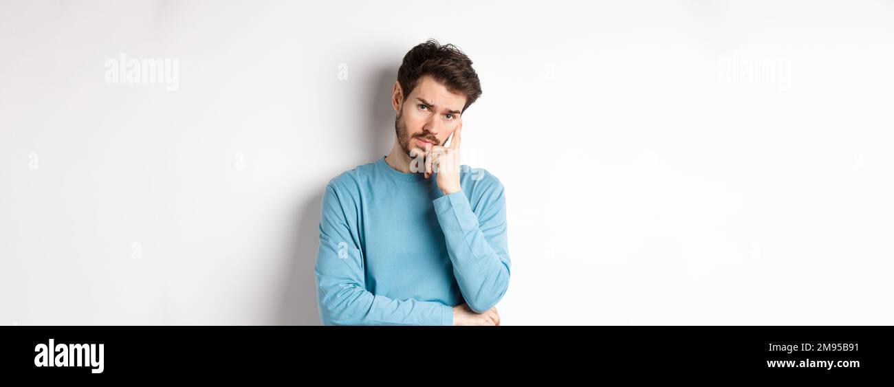 Skeptical young man listening to you with bored face, looking at camera ...