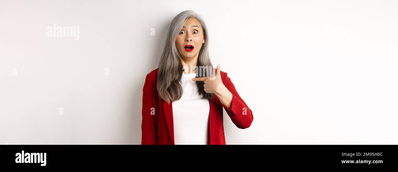 Surprised asian woman with grey hair, pointing at herself and gasping ...