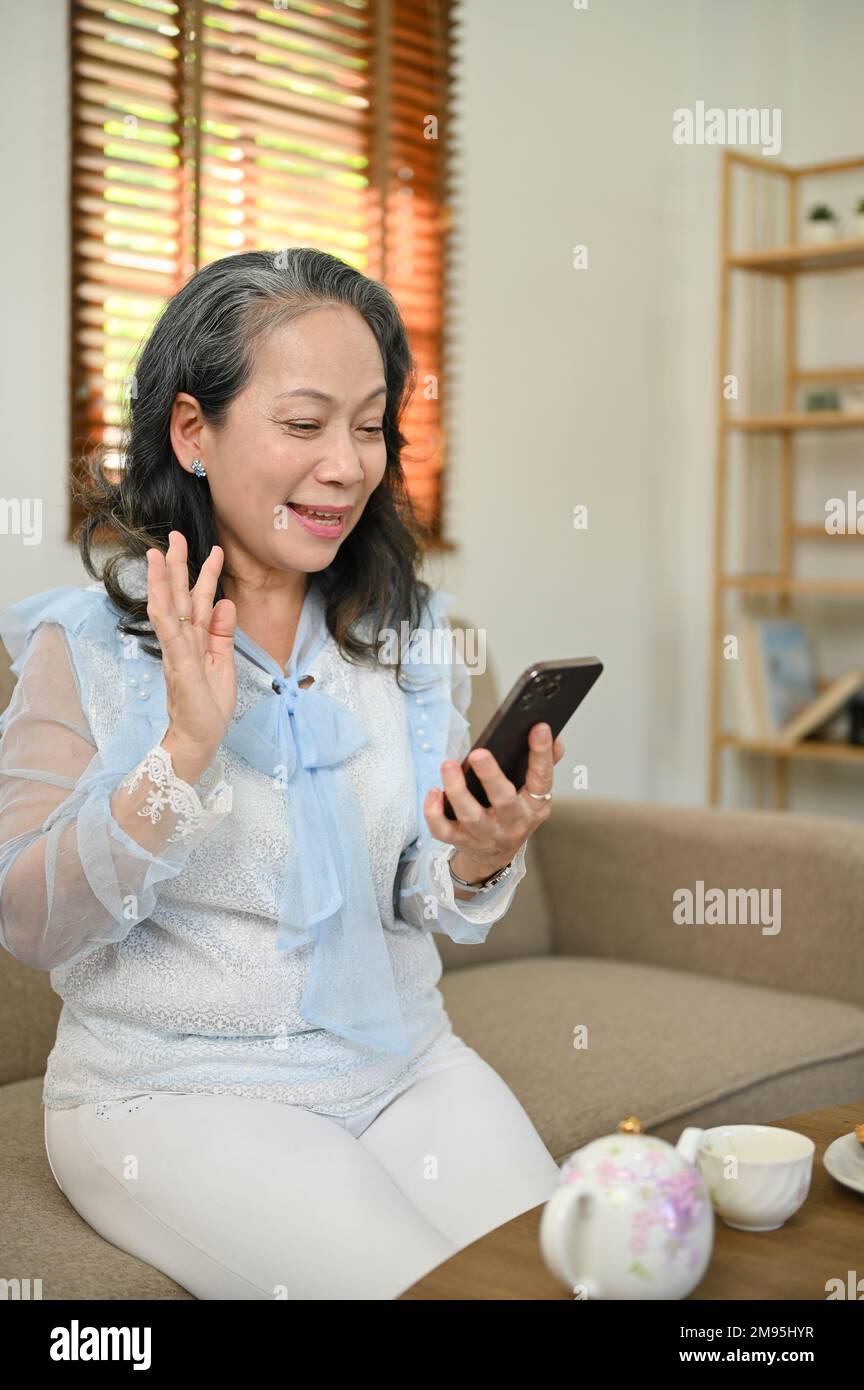 Happy and joyful 60s aged Asian woman is in the living room, waving ...
