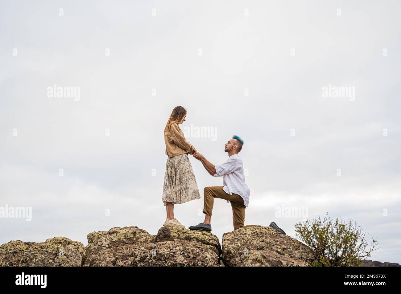 A young couple stands on a rocky cliff by the ocean, with the man down on one knee proposing to ...