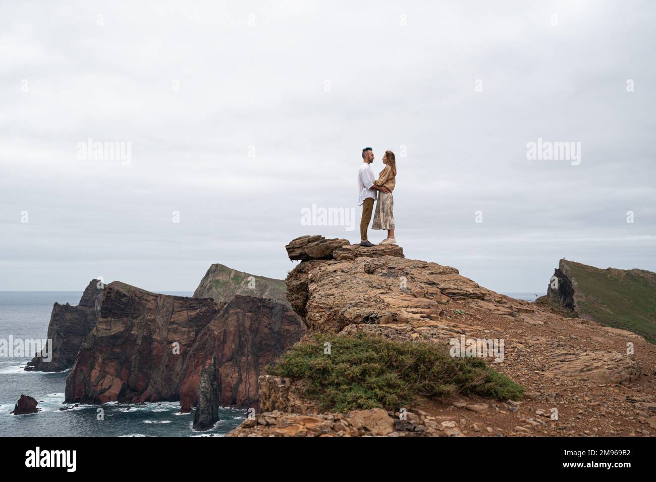 A young couple stands on a rocky cliff by the ocean, gazing into each other's eyes and holding ...