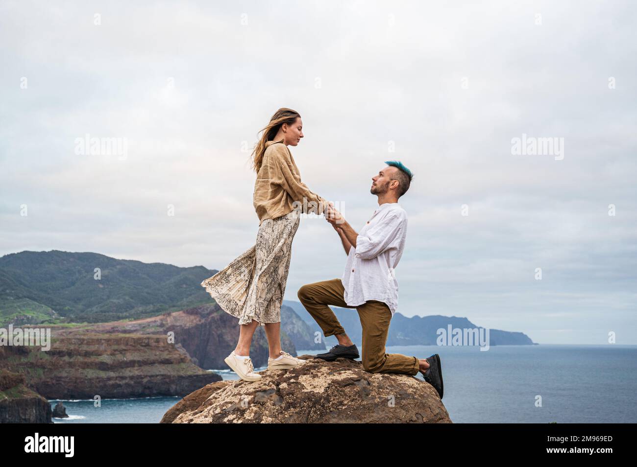 A young couple stands on a rocky cliff by the ocean, with the man down on one knee proposing to ...