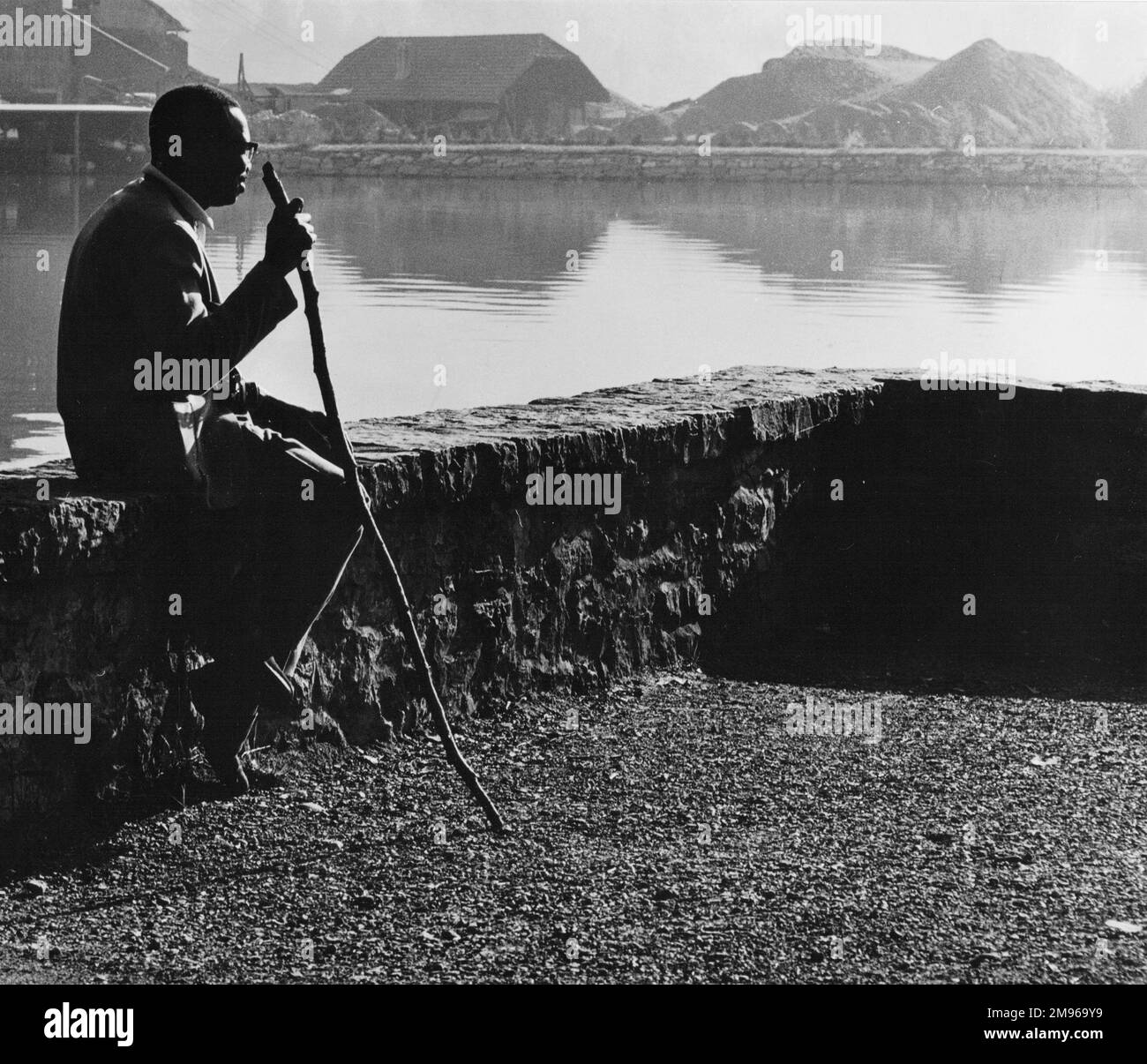 A man with a long stick sitting on a wall, looking out over a stretch ...