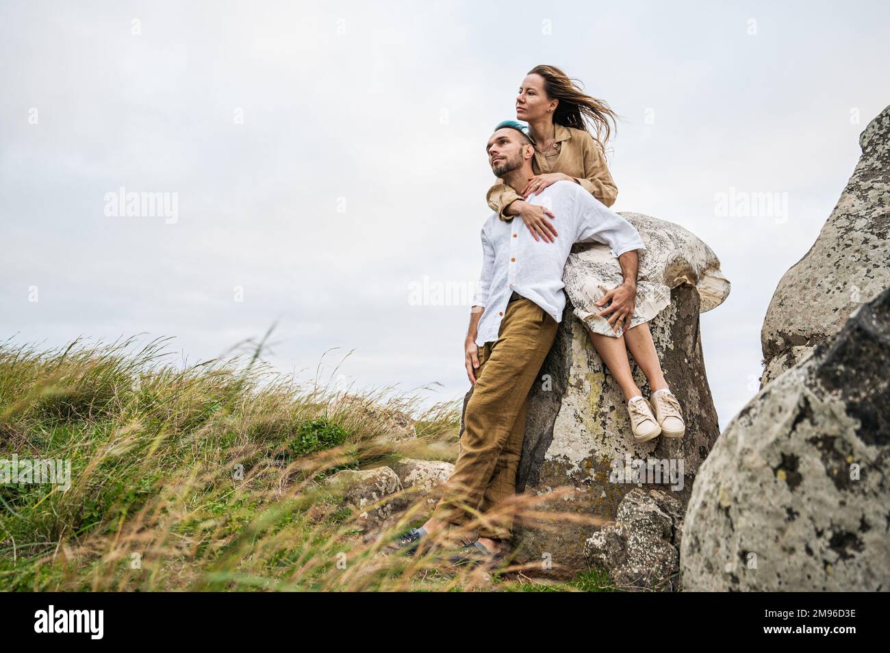 A young woman sits on a huge rock and embraces her partner from behind ...
