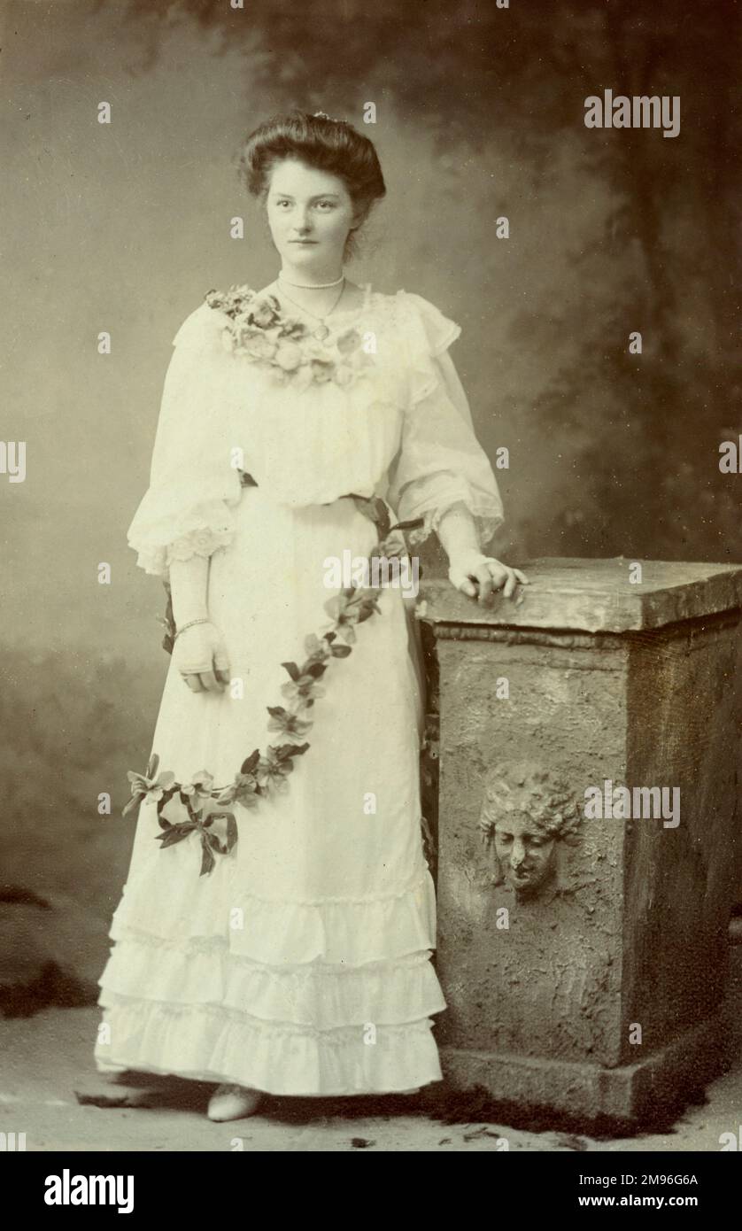 A woman stands by a stone plinth, dressed in a frilly white dress and ...