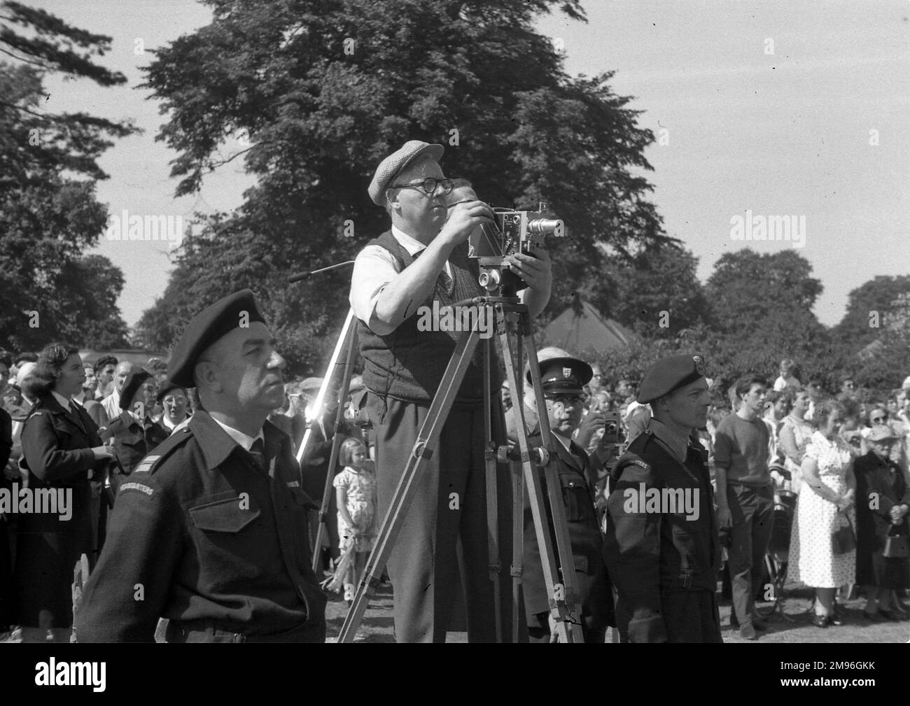 People in a park, watching an event. A man stands with a cine camera on ...