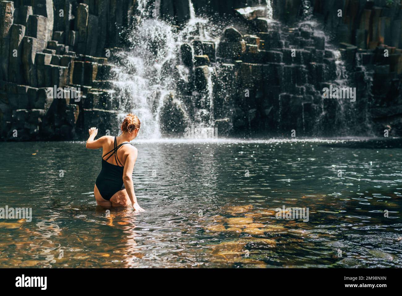 Caucasian woman in black swimsuit entering into waterfall lake ...