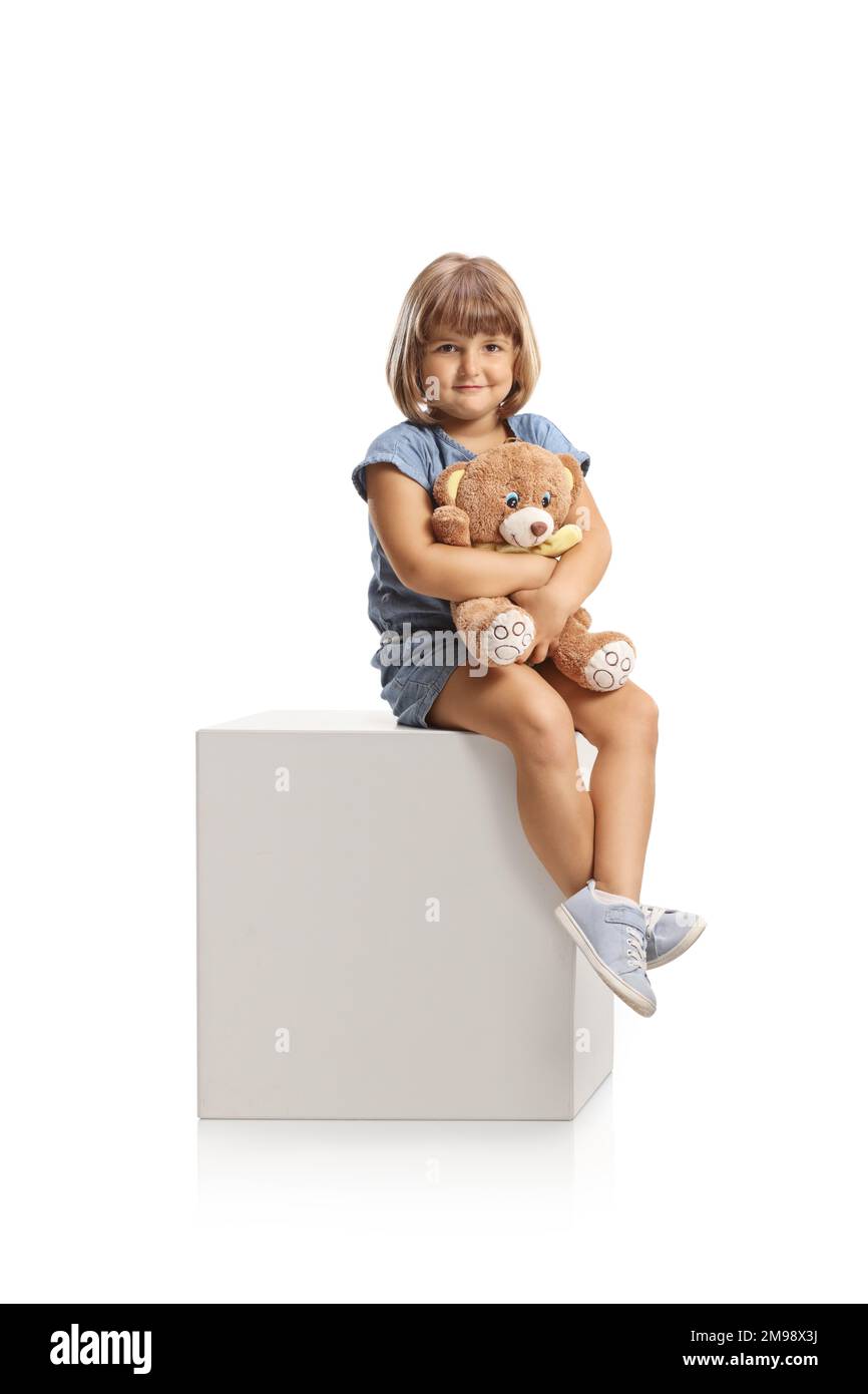 Cute little girl holding a teddy bear and sitting on a white cube ...