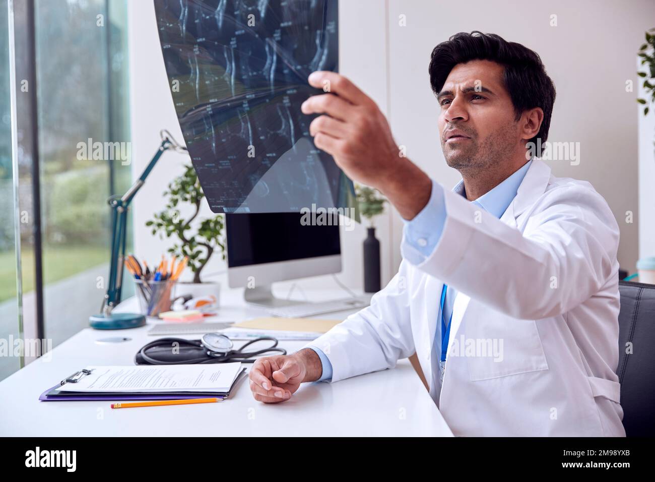 Male Doctor Wearing White Coat Sitting At Desk Looking At CT Or MRI ...