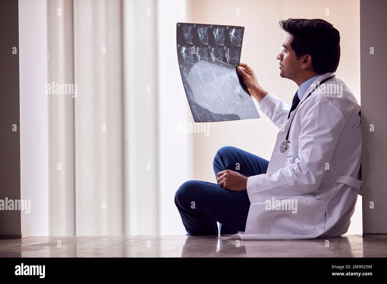 Male Doctor Wearing White Coat Sitting On Floor In Hospital Corridor ...