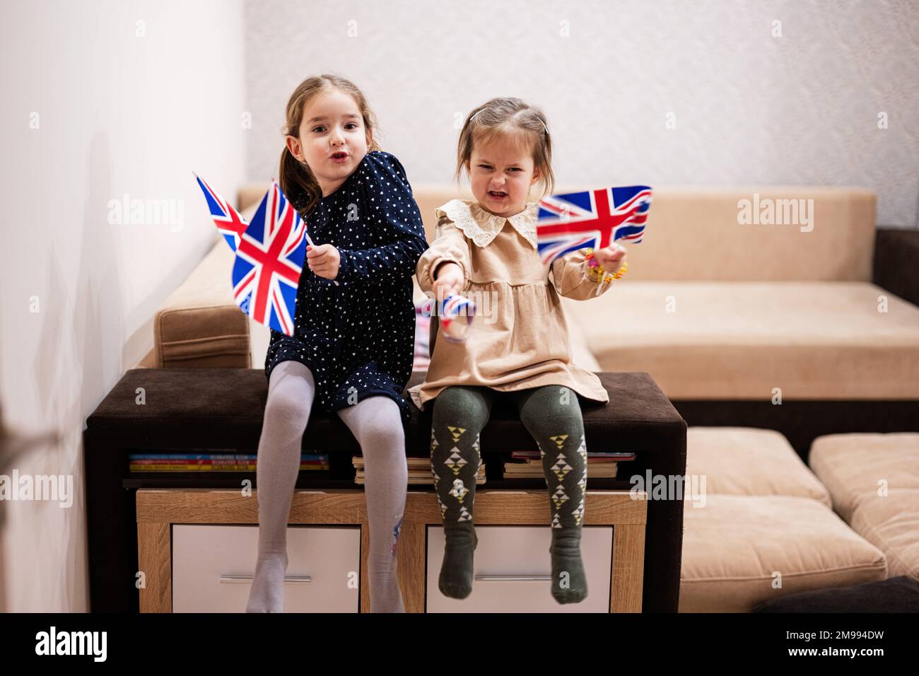 Two sisters are sitting on a couch at home with british flags on hands ...