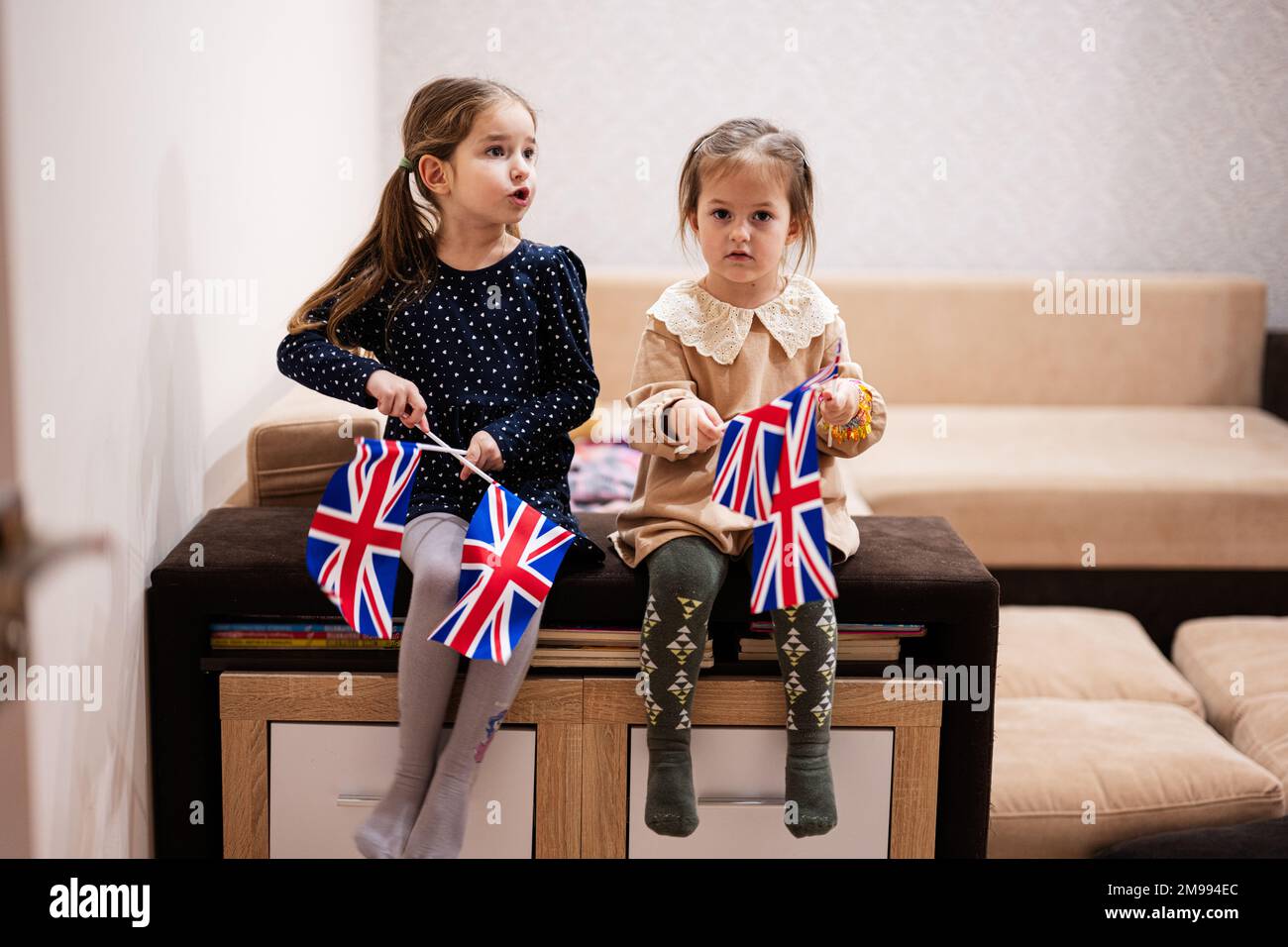 Two sisters are sitting on a couch at home with british flags on hands ...