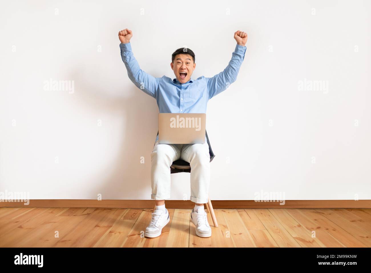 Overjoyed asian man making YES gesture, sitting on chair with laptop ...