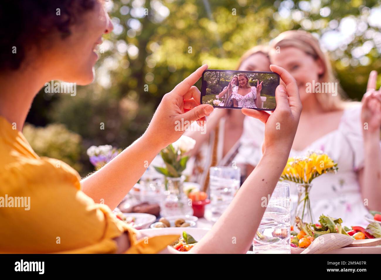 Woman Taking Photo Of Female Friends Sitting Outdoors In Garden At Home ...