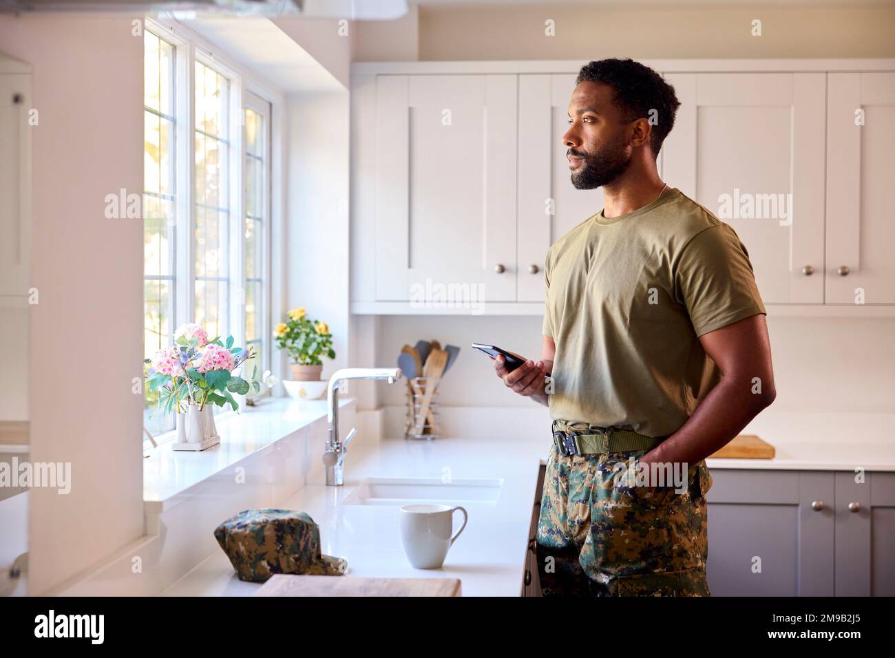 Thoughtful Male Soldier In Uniform With Mobile Phone In Kitchen On Home ...