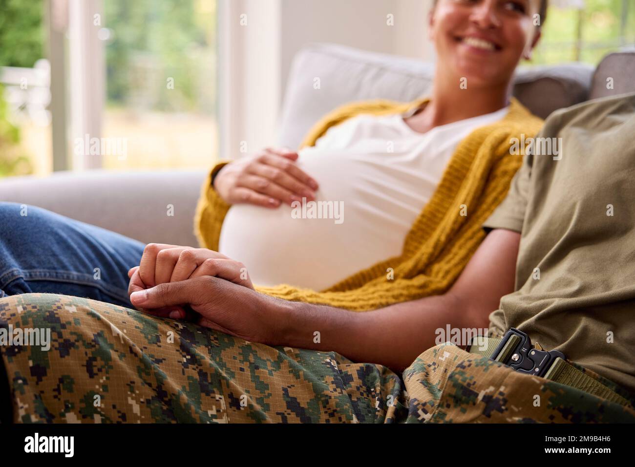 Pregnant Couple With Woman Sitting Next To Army Husband In Uniform Holding Hands Home On Leave