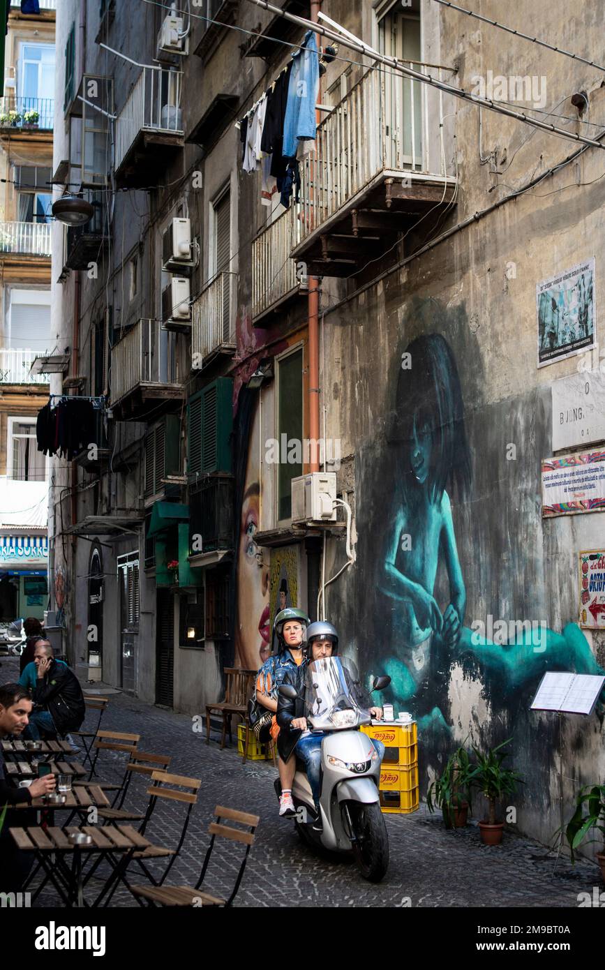 A couple ride a scooter through an alley in Naples, there is a large ...