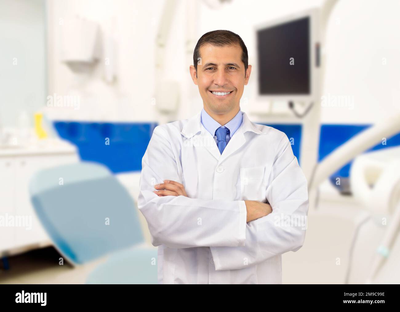 A male dentist standing next to his desk with his arms folded and ...