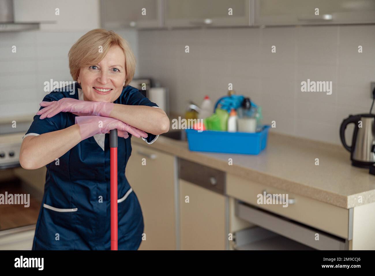 Portrait of smiling senior housekeeping standing with mop on kitchen ...